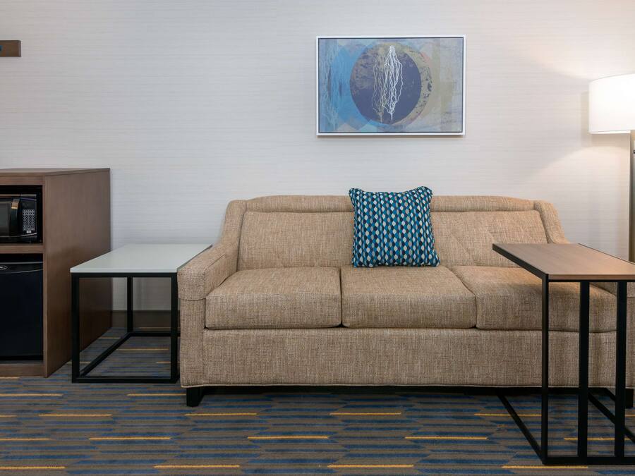 Quarto de hotel com um sofá bege, uma almofada estampada, uma mesinha, um micro-ondas e uma luminária.