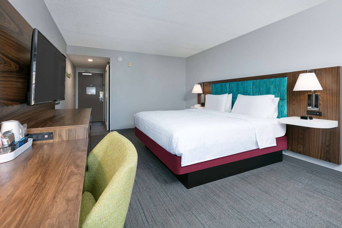 Quarto de hotel com cama, escrivaninha com televisão, cadeira e mesa de cabeceira com luminárias.