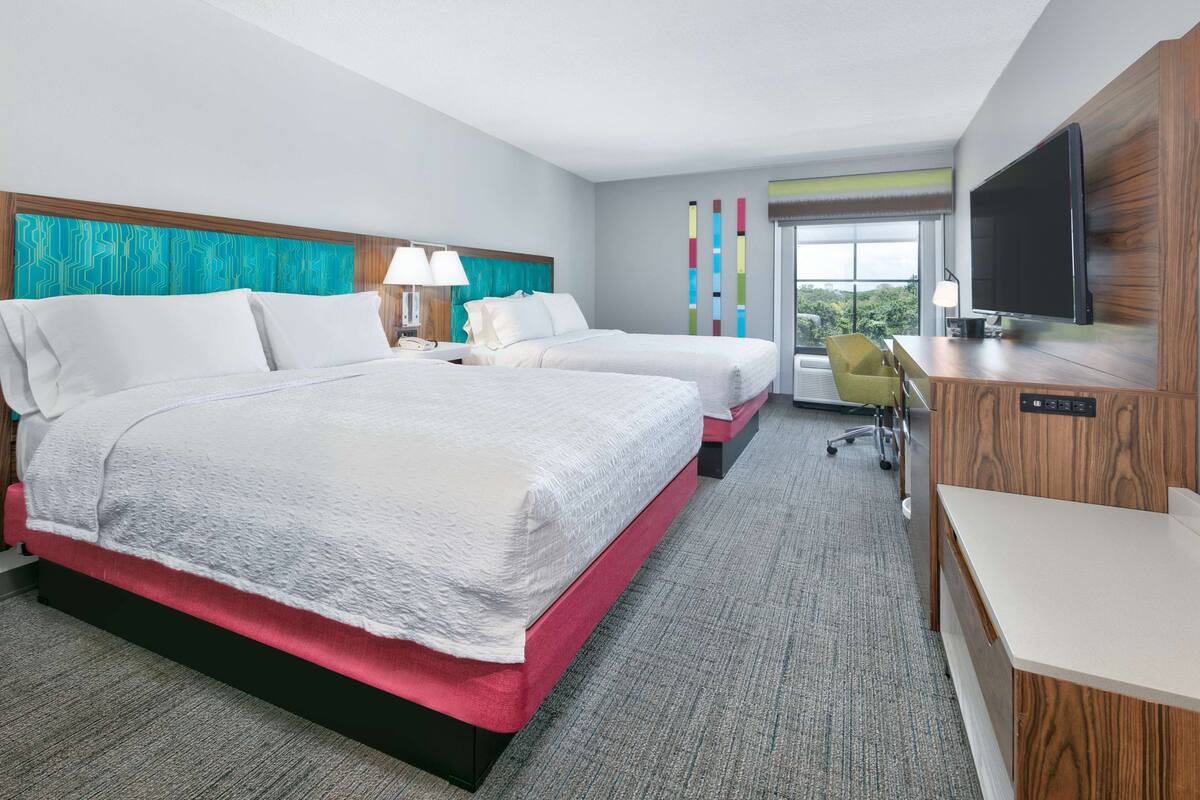 Quarto de hotel com duas camas, uma escrivaninha, uma televisão e uma janela com vista.