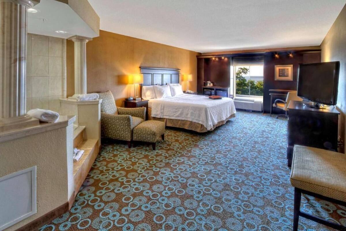 Quarto de hotel com uma cama, duas cadeiras, uma escrivaninha, uma televisão e um tapete estampado.