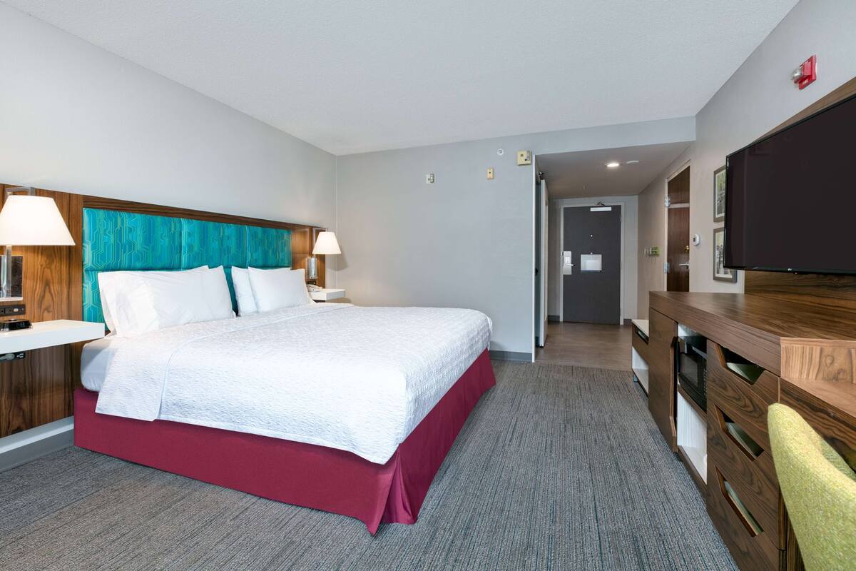 Quarto de hotel com cama grande, escrivaninha, cadeira, televisão de tela plana e porta para outro cômodo.