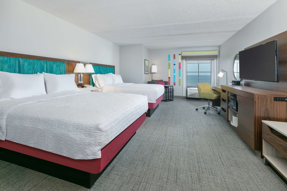 Quarto de hotel com duas camas, uma escrivaninha, televisão e uma janela grande.
