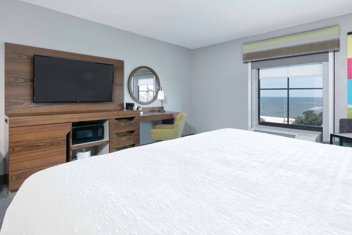 Quarto de hotel com uma cama grande, uma TV fixada num móvel de madeira, uma escrivaninha com uma cadeira, uma janela com vista para o mar e uma pequena mesa com um abajur.