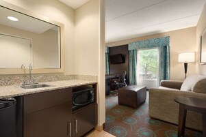 Quarto de hotel com cozinha compacta, televisão de tela plana, sofá e janela com cortinas.