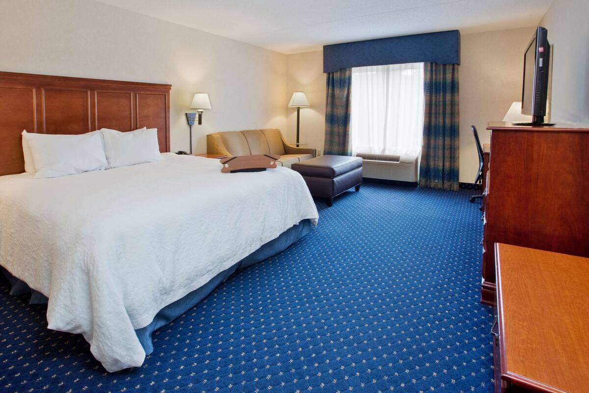 Quarto de hotel com uma cama grande, uma escrivaninha, uma cadeira, televisão e janela com cortinas.