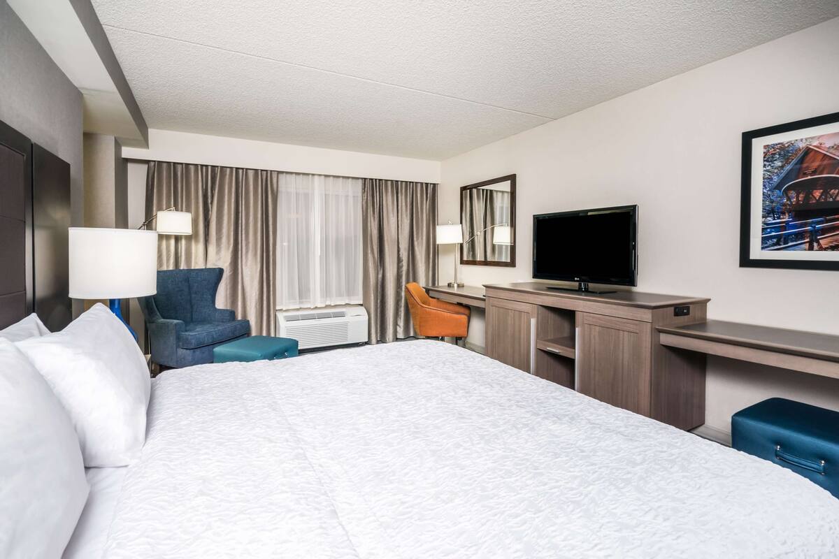 Quarto de hotel com uma cama grande, uma escrivaninha, televisão de tela plana, uma cadeira e uma janela com cortinas.