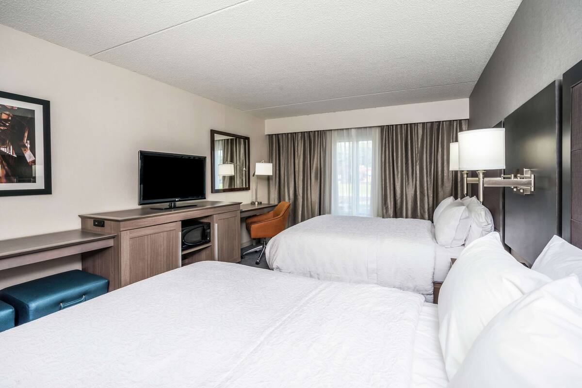 Quarto de hotel com duas camas, uma escrivaninha com computador, televisão e uma cadeira.