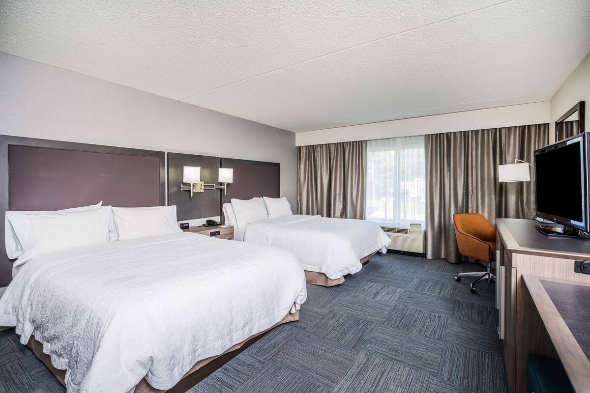 Quarto de hotel com duas camas, uma escrivaninha, uma cadeira, televisão e janela com cortinas.