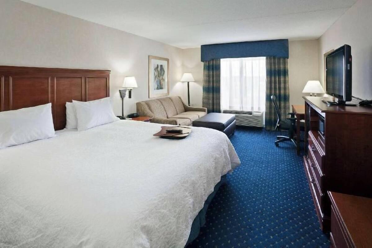 Quarto de hotel com uma cama grande, uma escrivaninha com computador, televisão e um sofá.