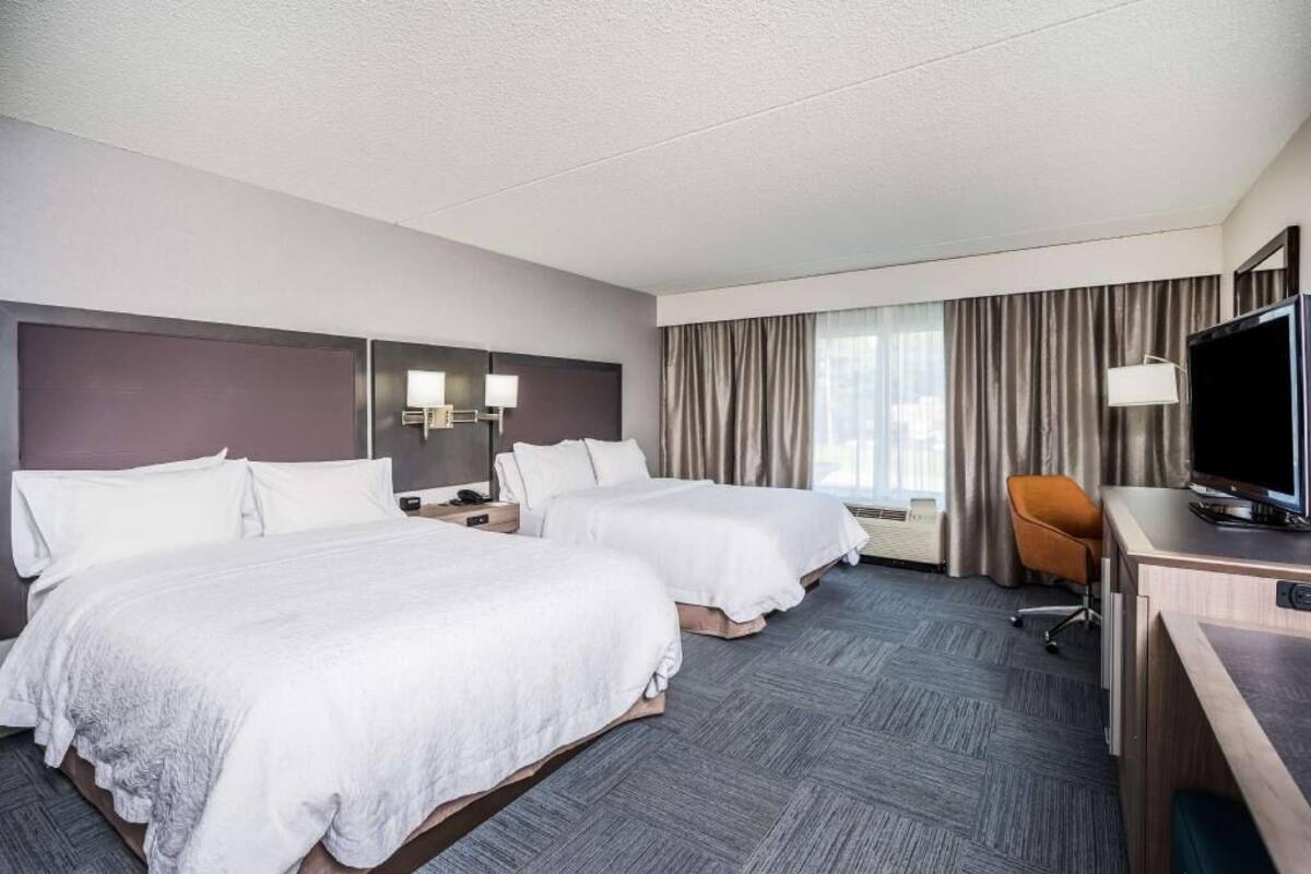 Quarto de hotel com duas camas, uma escrivaninha, televisão e janela com cortinas.