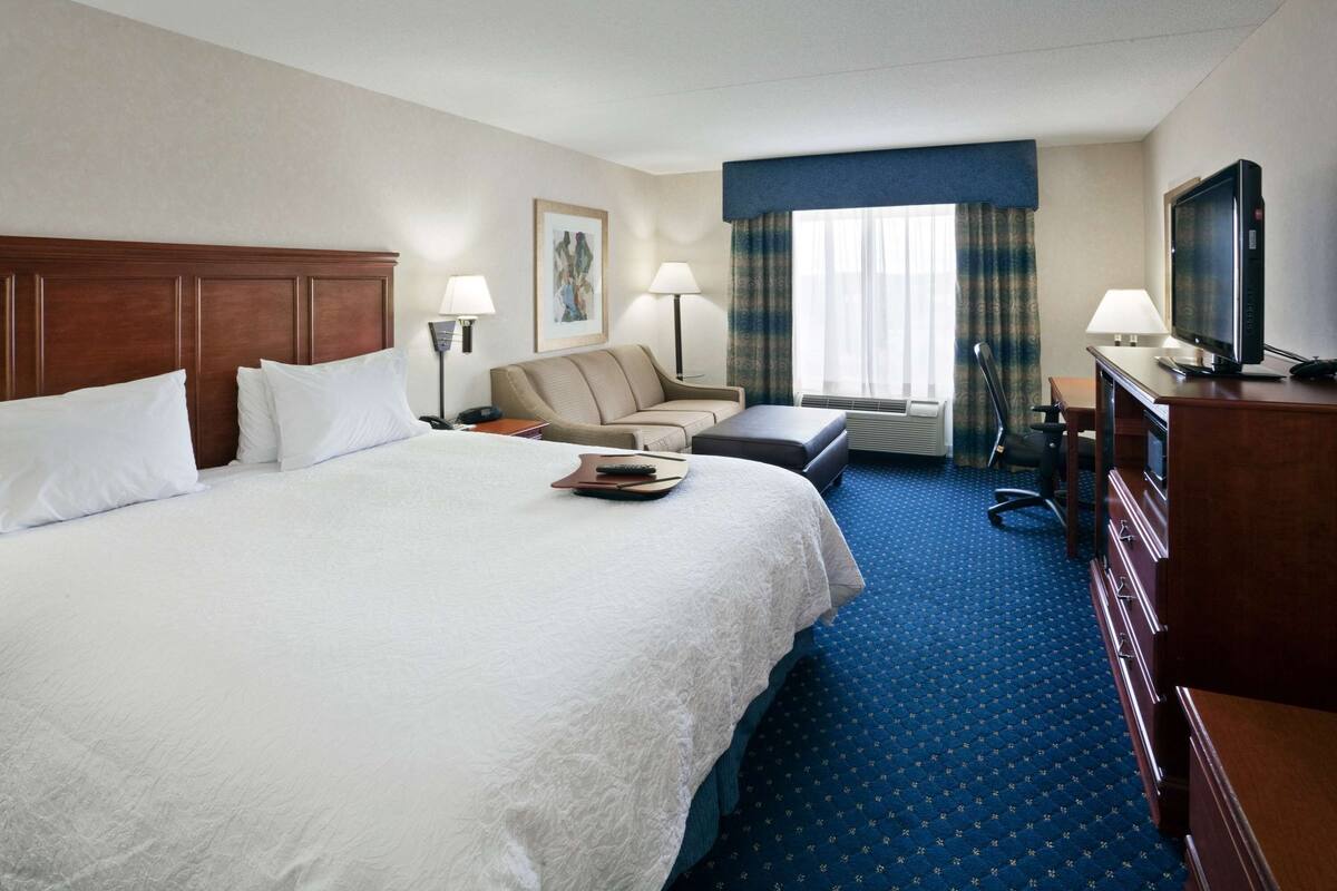 Quarto de hotel com uma cama grande, uma escrivaninha, uma cadeira, uma televisão e uma cômoda.