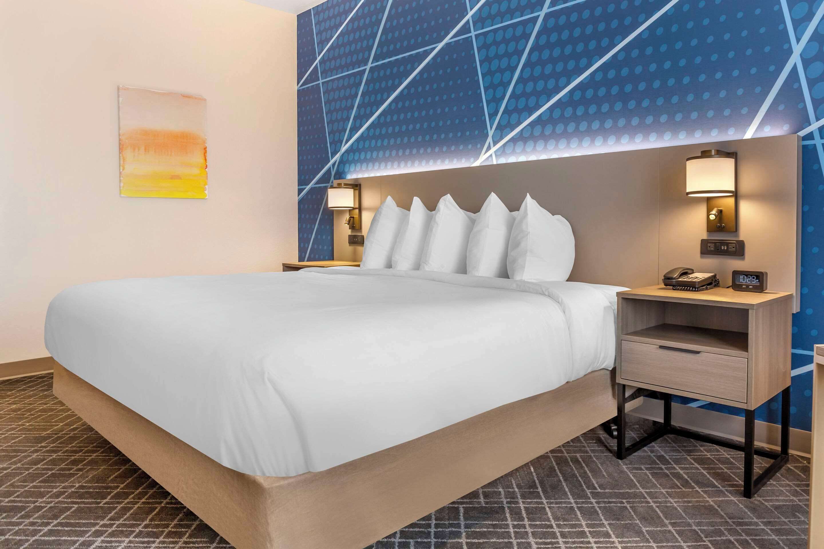 Quarto de hotel com uma cama grande, uma mesa de cabeceira com abajur e uma parede com padrão geométrico.