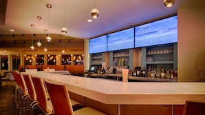 Uma área de bar com um balcão comprido, várias luminárias pendentes e uma tela grande exibindo uma vista urbana.