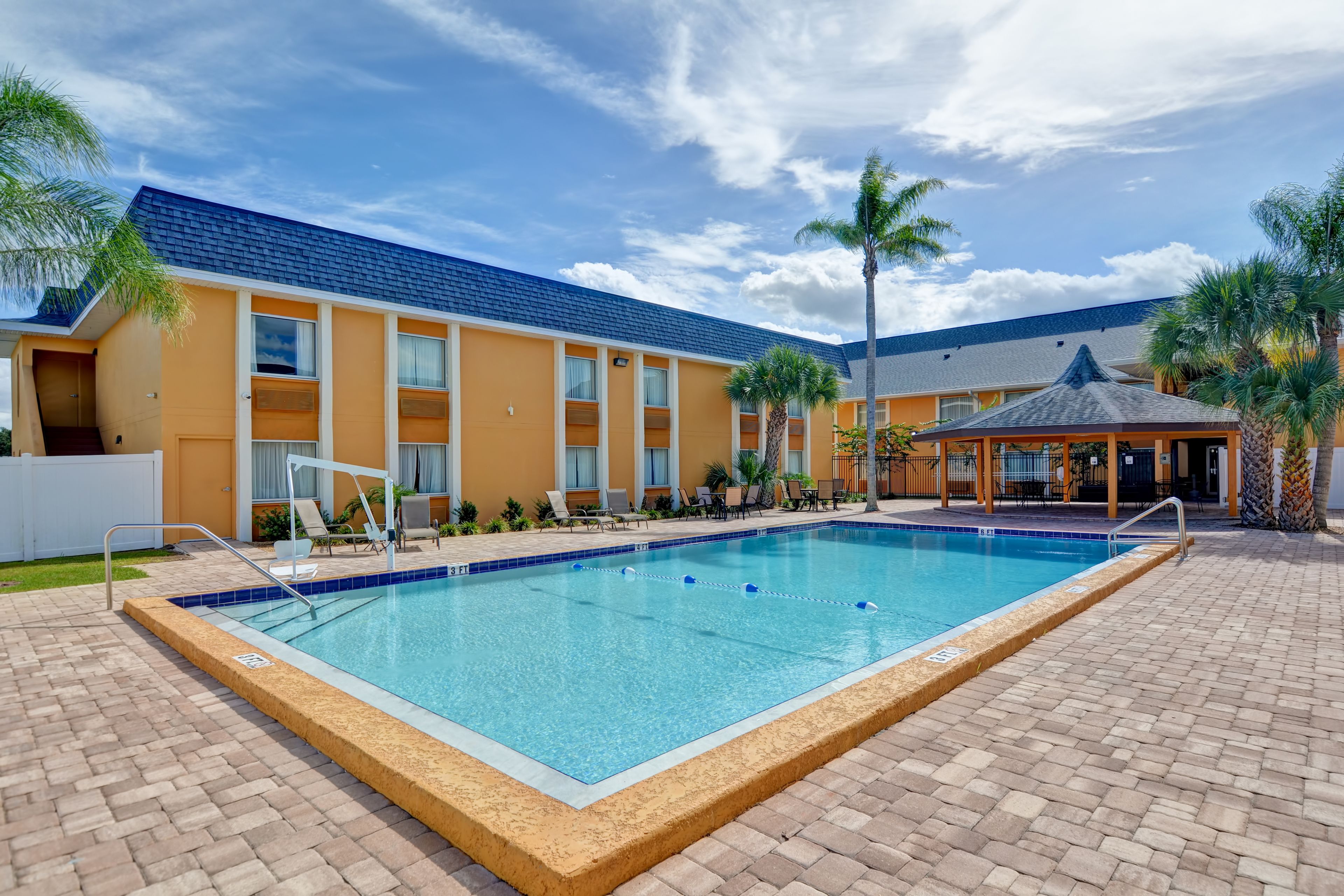 Piscina com deck revestido, área de estar coberta e edifício com telhado azul e paredes amarelas.