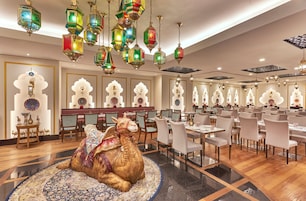 Uma sala de jantar espaçosa com várias mesas e cadeiras, luminárias pendentes ornamentadas e uma estátua de camelo.