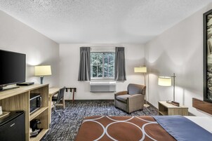 Quarto de hotel com cama, escrivaninha, cadeira, televisão, ar condicionado e janela com cortinas.