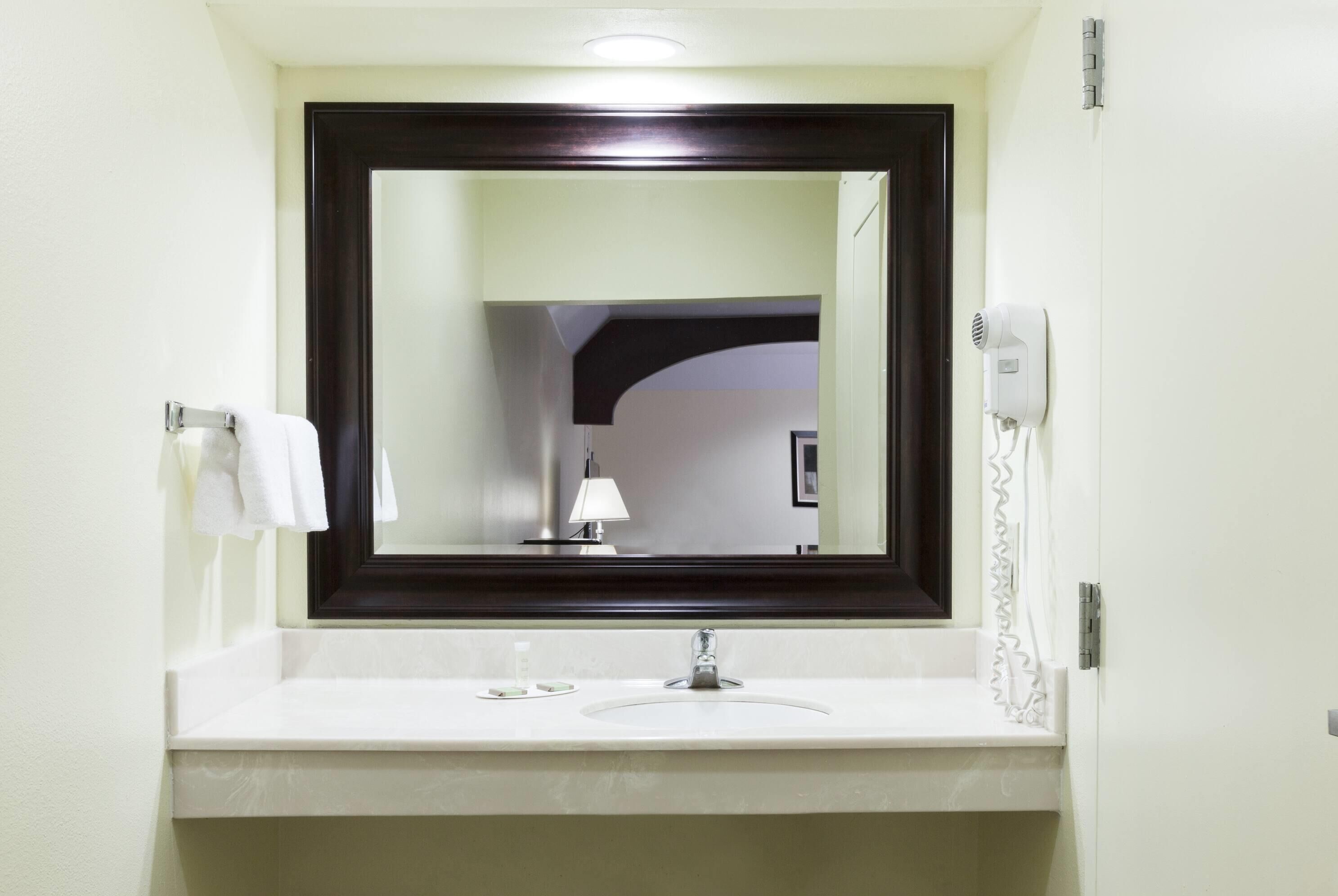 Casa de banho com uma pia branca, um espelho com moldura escura, um porta-toalhas e um secador de cabelo preso à parede.