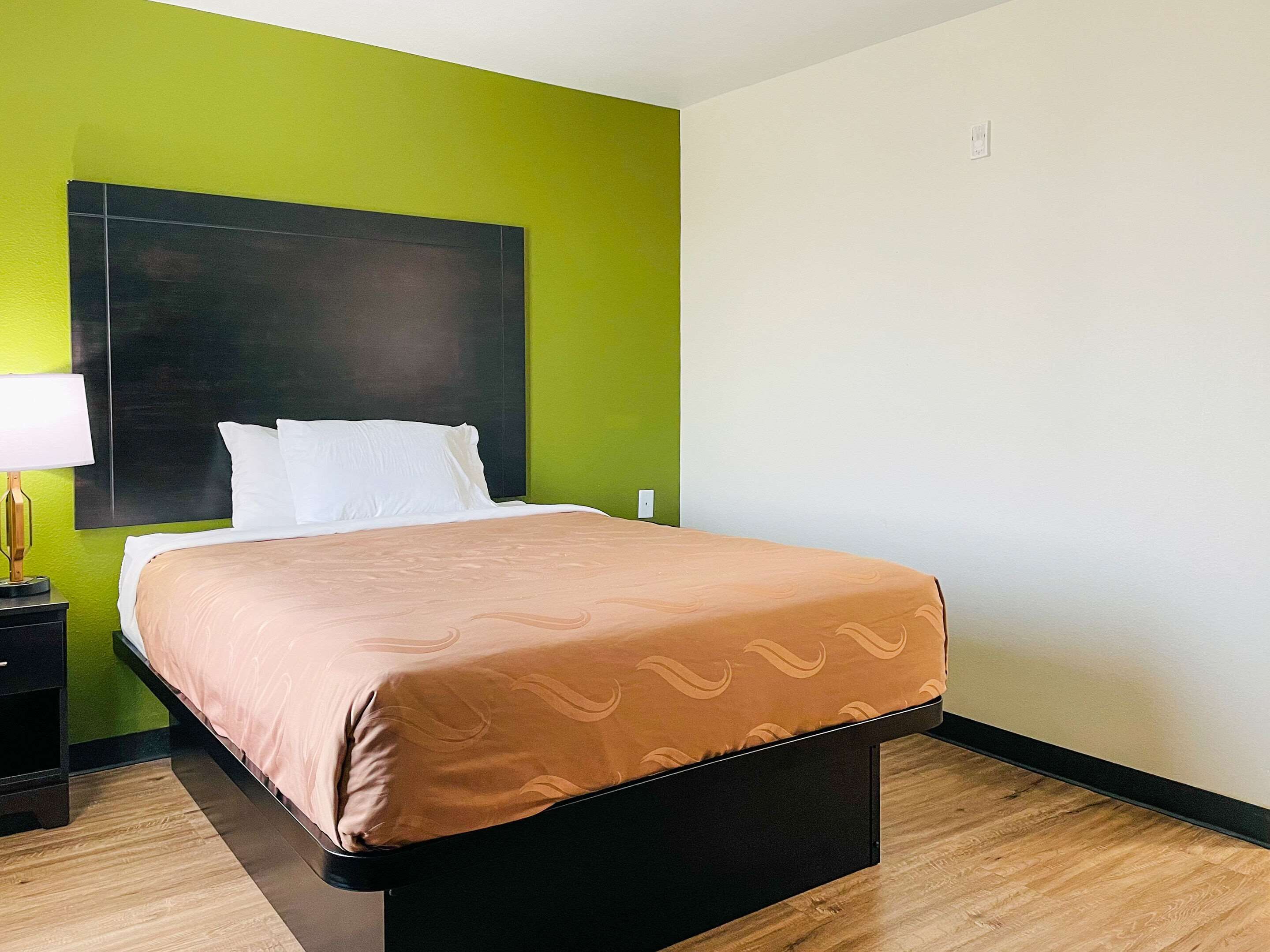 Quarto de hotel com uma cama, uma mesinha de cabeceira e uma luminária.