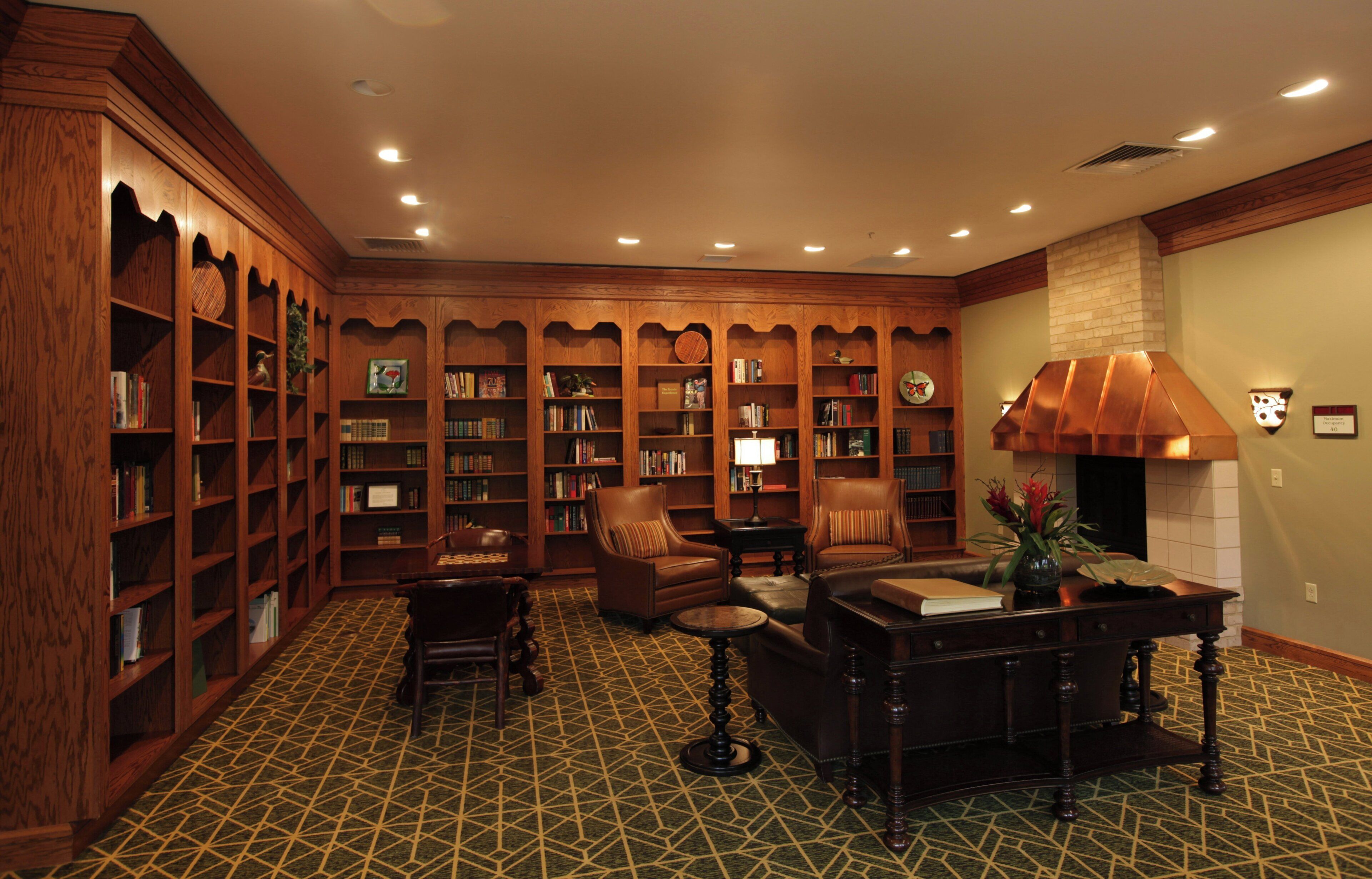 Uma biblioteca com estantes de madeira, poltronas de couro, lareira e tapete estampado.