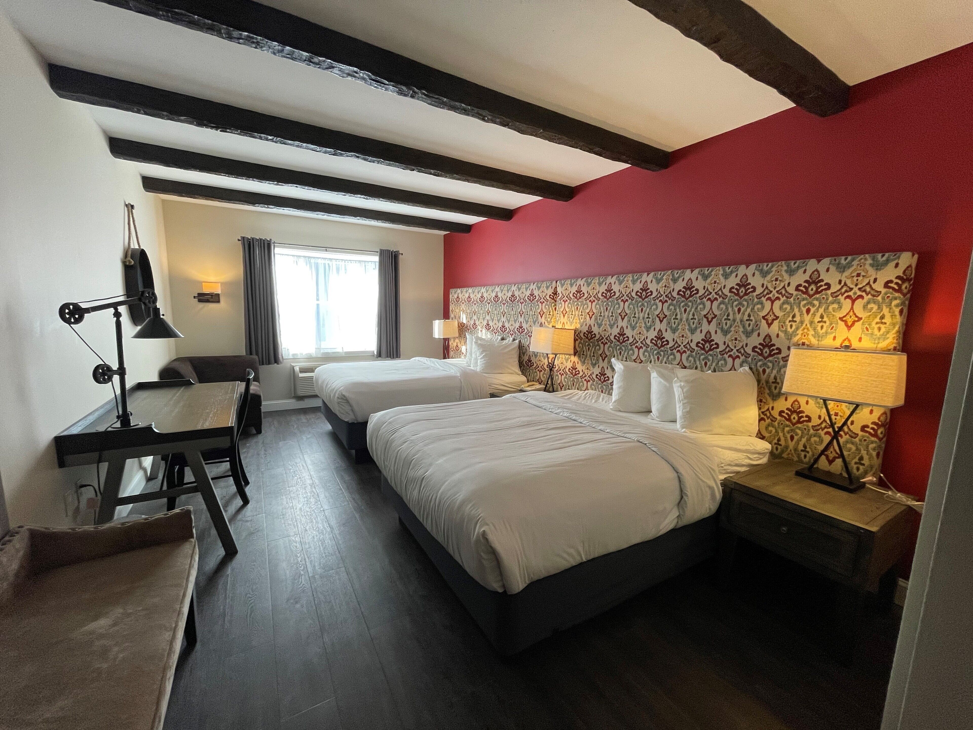 Quarto de hotel com duas camas, uma escrivaninha, uma cadeira, luminária de mesa e cabeceira estampada.