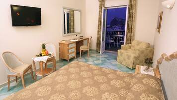 Quarto de hotel com cama, escrivaninha, cadeira, sofá, televisão, janela com cortinas e varanda com cadeiras.