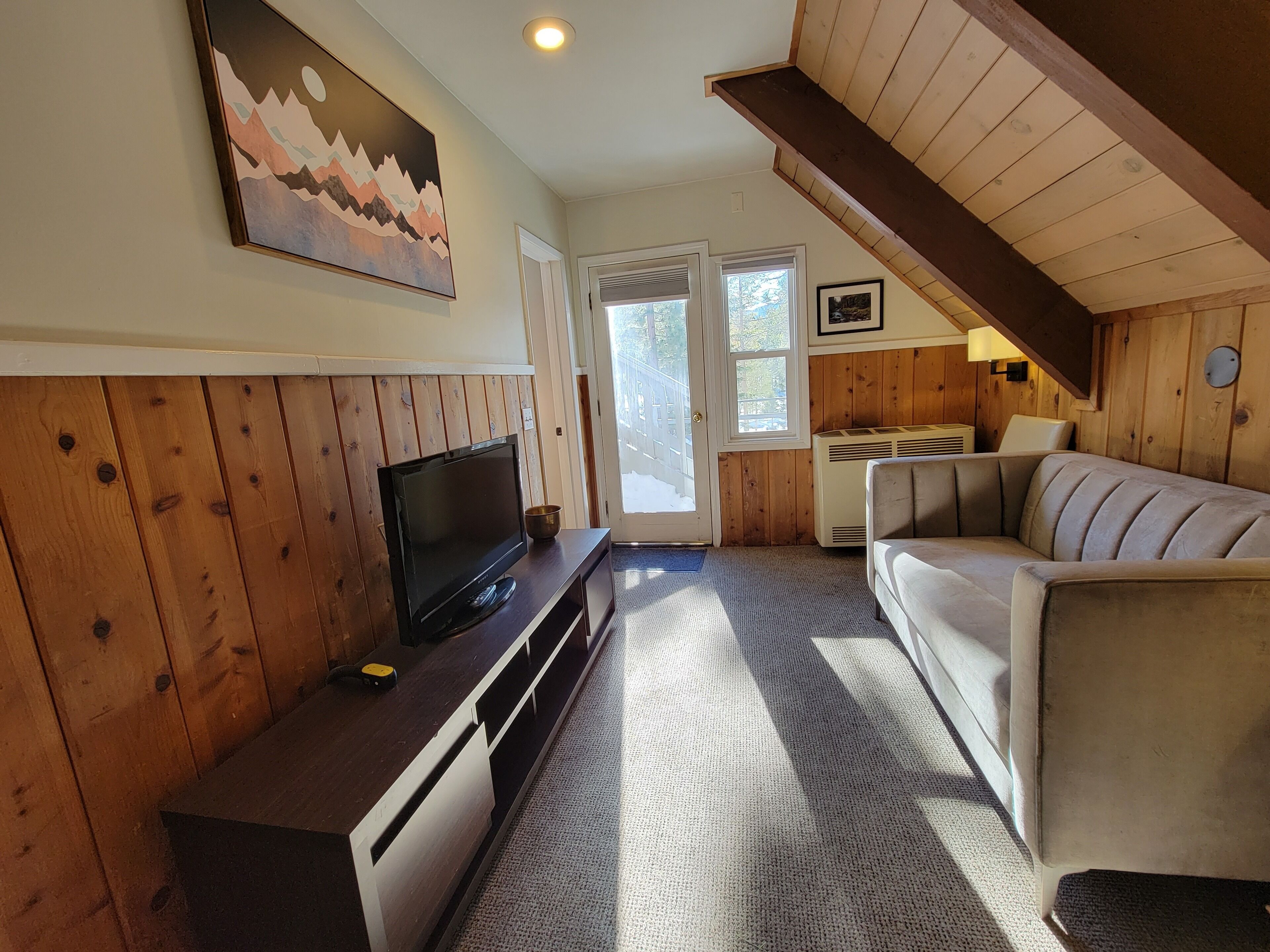 Uma sala de estar aconchegante com televisão, paredes de madeira, um sofá e uma porta que dá para uma cozinha.
