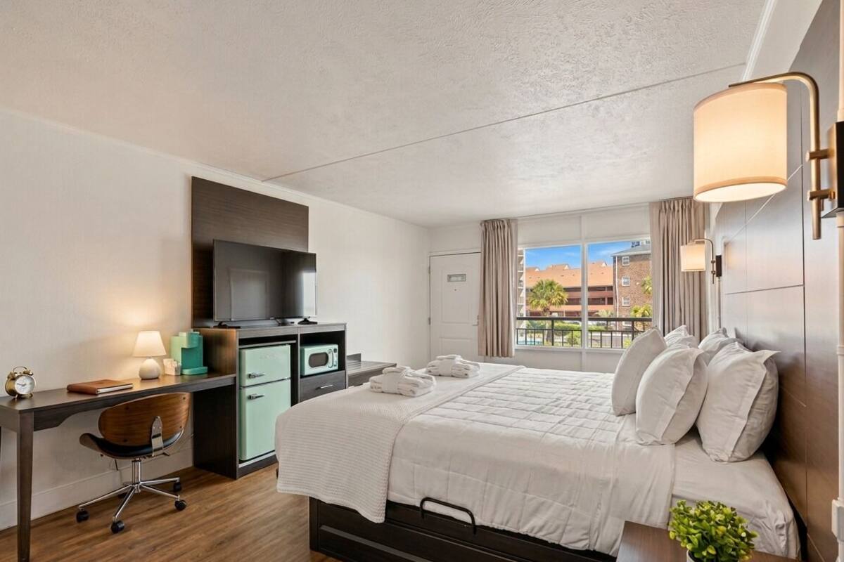 Quarto de hotel com cama, escrivaninha, televisão e janela com vista para edifícios.