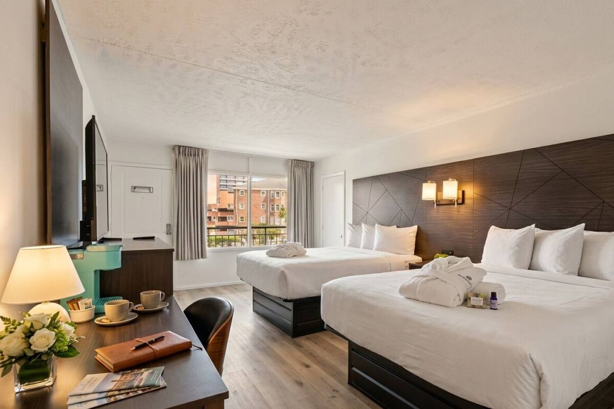 Quarto de hotel com duas camas, uma escrivaninha com luminária, televisão e vista para edifícios.