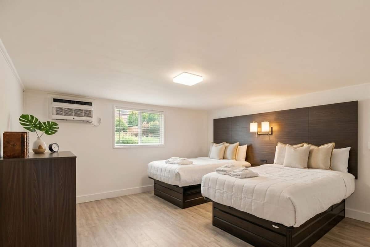 Quarto com duas camas, uma escrivaninha, uma planta e uma janela.