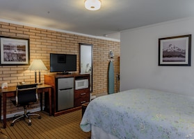 Quarto de hotel com parede de tijolos, televisão de tela plana, geladeira, micro-ondas, escrivaninha com cadeira, cama com colcha florida e duas fotos emolduradas.