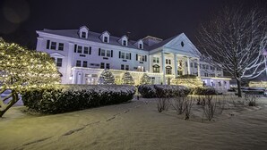 Um edifício grande e branco, com várias janelas e uma entrada imponente, rodeado por paisagismo coberto de neve e iluminado por luzes artificiais.