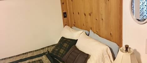 Quarto com cabeceira de madeira, cama com várias almofadas, manta xadrez, abajur e quadro na parede.