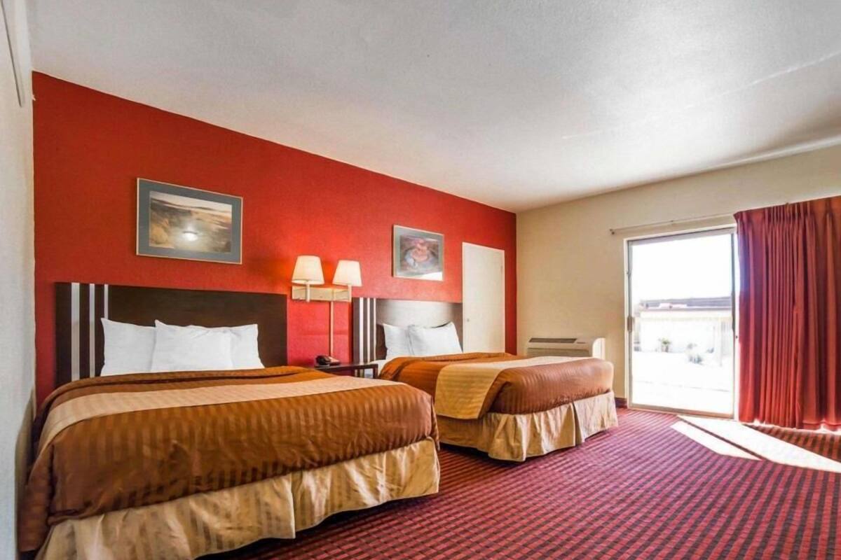 Quarto de hotel com duas camas, um tapete vermelho, uma janela grande com cortinas e um quadro na parede.
