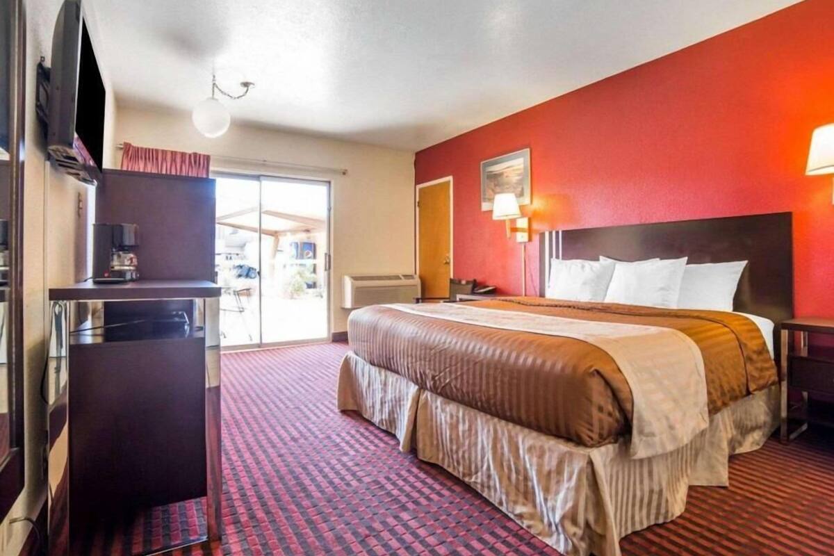 Quarto de hotel com cama, secretária com computador, televisão e porta de correr para o exterior.