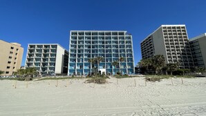 Um hotel à beira-mar com vários andares, varandas e uma praia de areia em primeiro plano.