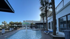 Uma área de piscina com espreguiçadeiras, um elemento aquático, palmeiras e um edifício moderno ao fundo.