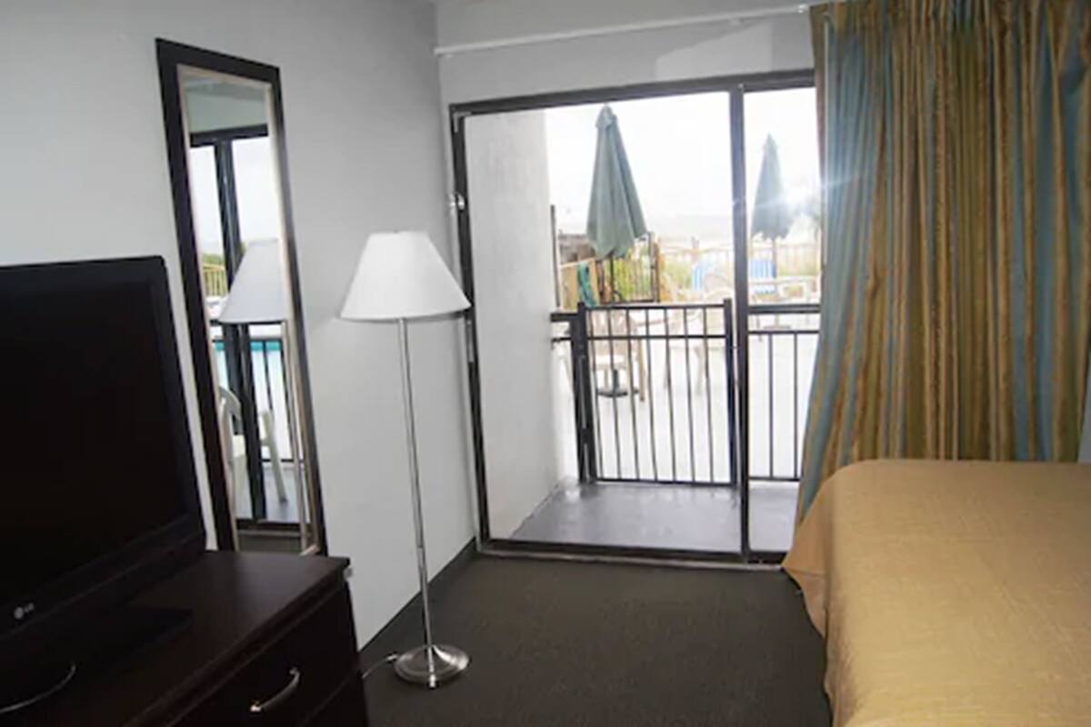 Quarto de hotel com cama, televisão em uma cômoda, luminária e varanda com vista para o exterior.