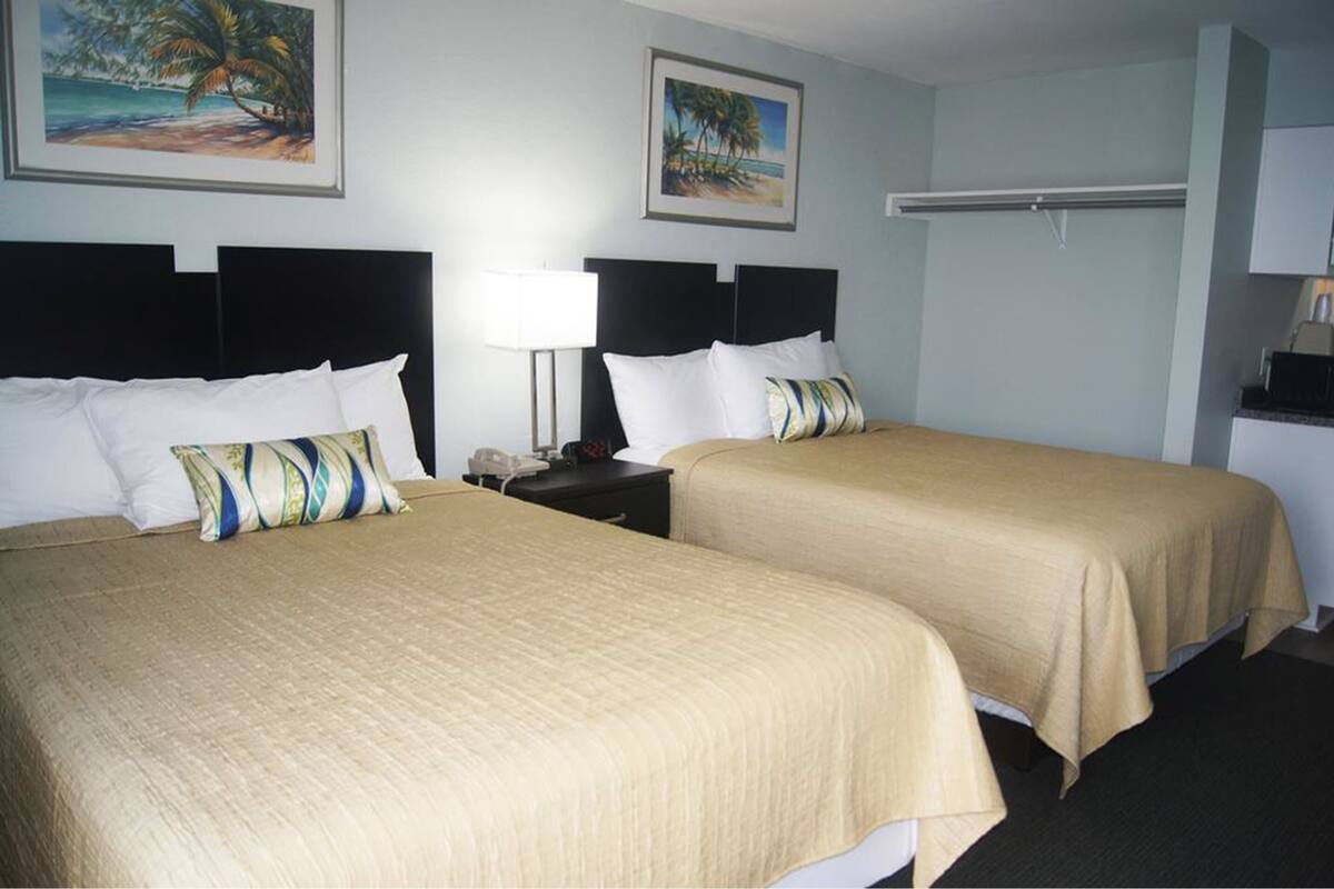Quarto de hotel com duas camas, cada uma com colcha e almofadas bege, uma mesa de cabeceira com abajur, e um quadro com uma cena de praia na parede.