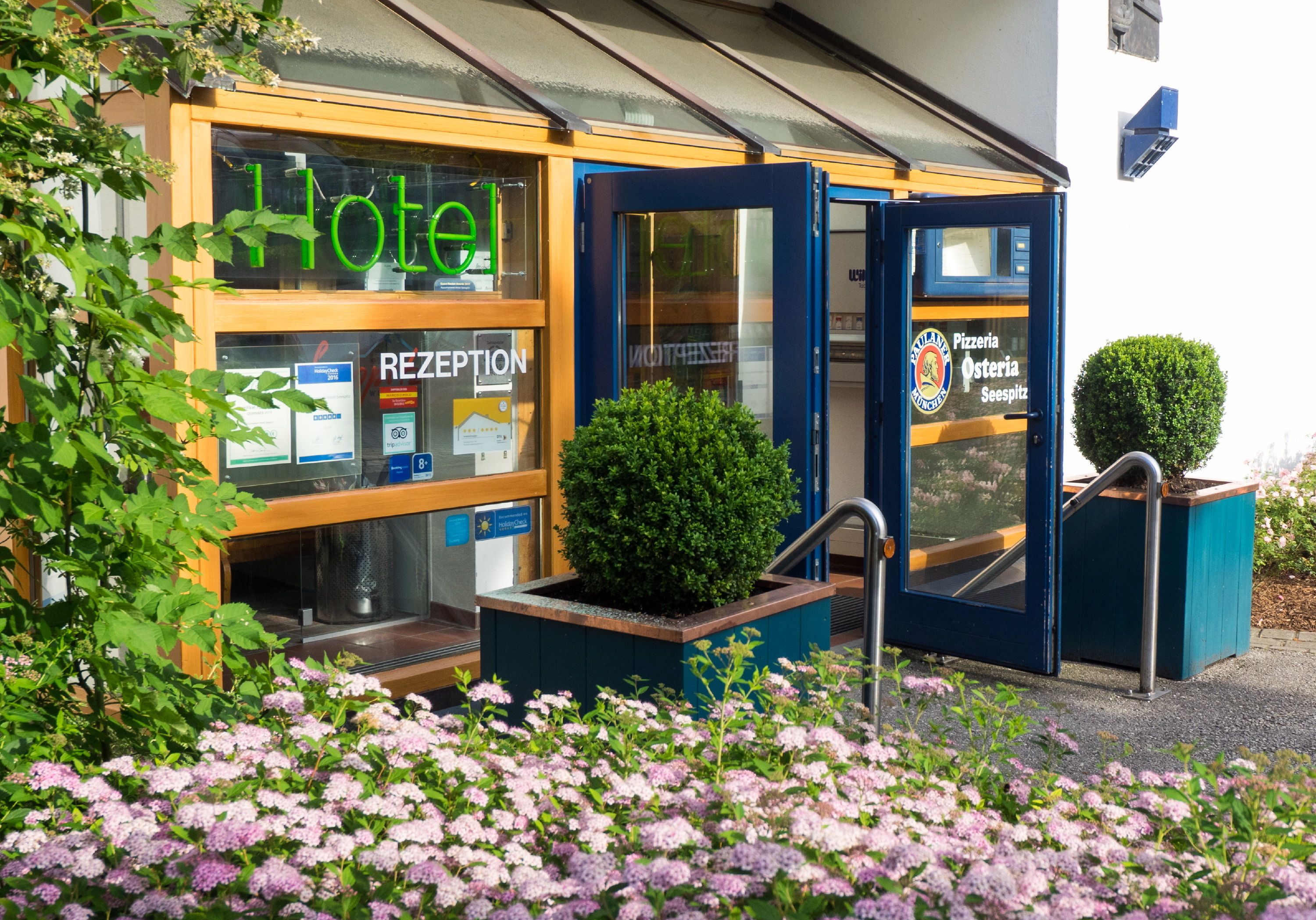 Entrada de hotel com balcão de recepção, placa de pizzaria e um pequeno jardim com flores.
