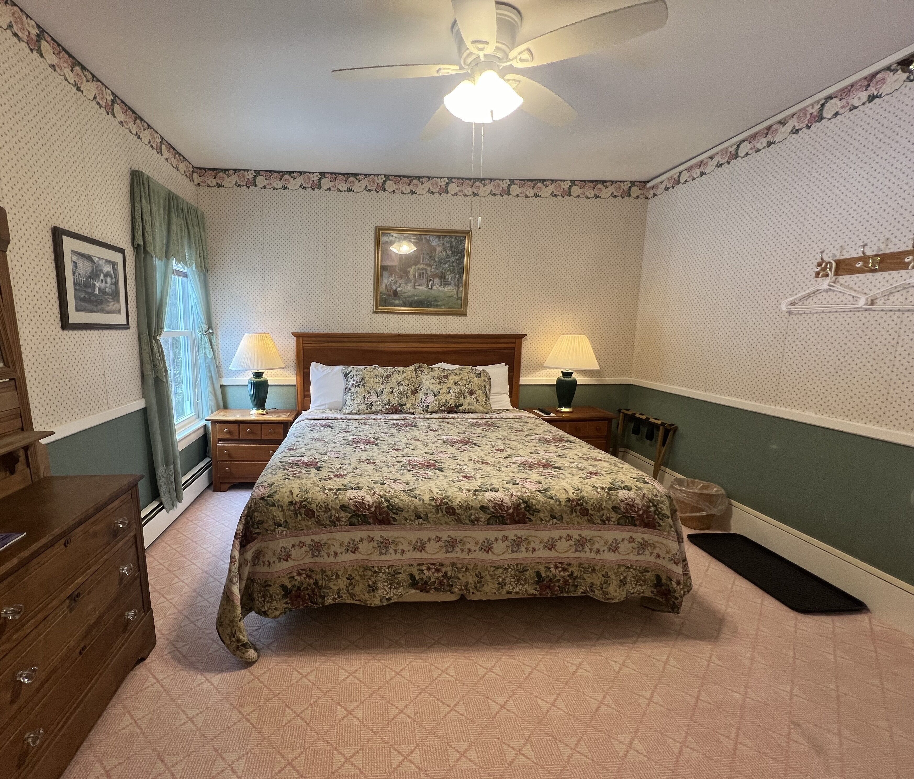 Quarto com cama, duas mesas de cabeceira, cómoda, ventoinha no teto, quadro na parede e janela com cortinas.