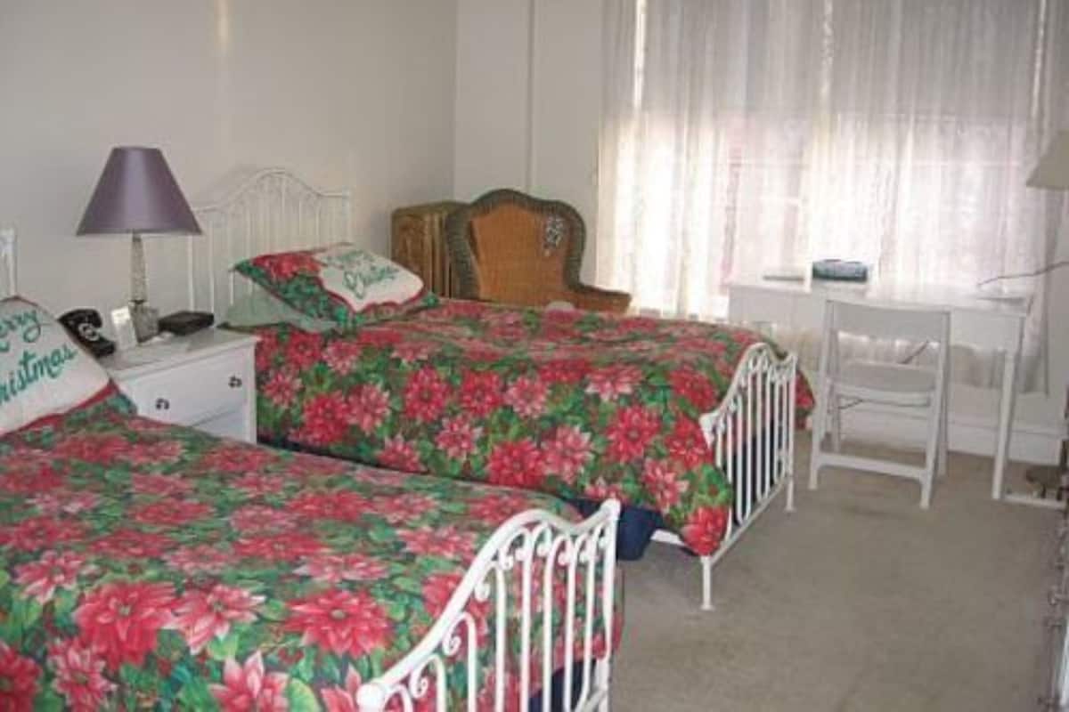 Quarto com duas camas, uma mesa de cabeceira, uma luminária, uma cadeira e uma escrivaninha.