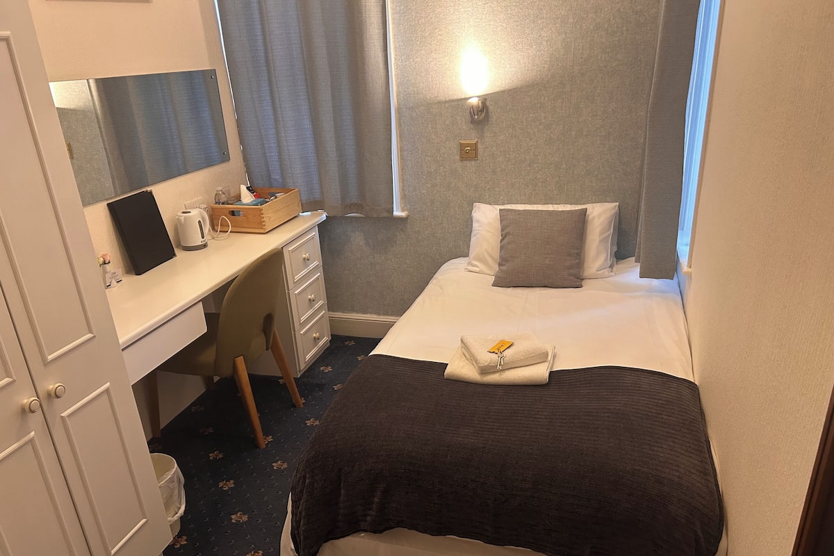 Quarto de hotel com cama, escrivaninha, cadeira, cômoda e janela com cortinas.