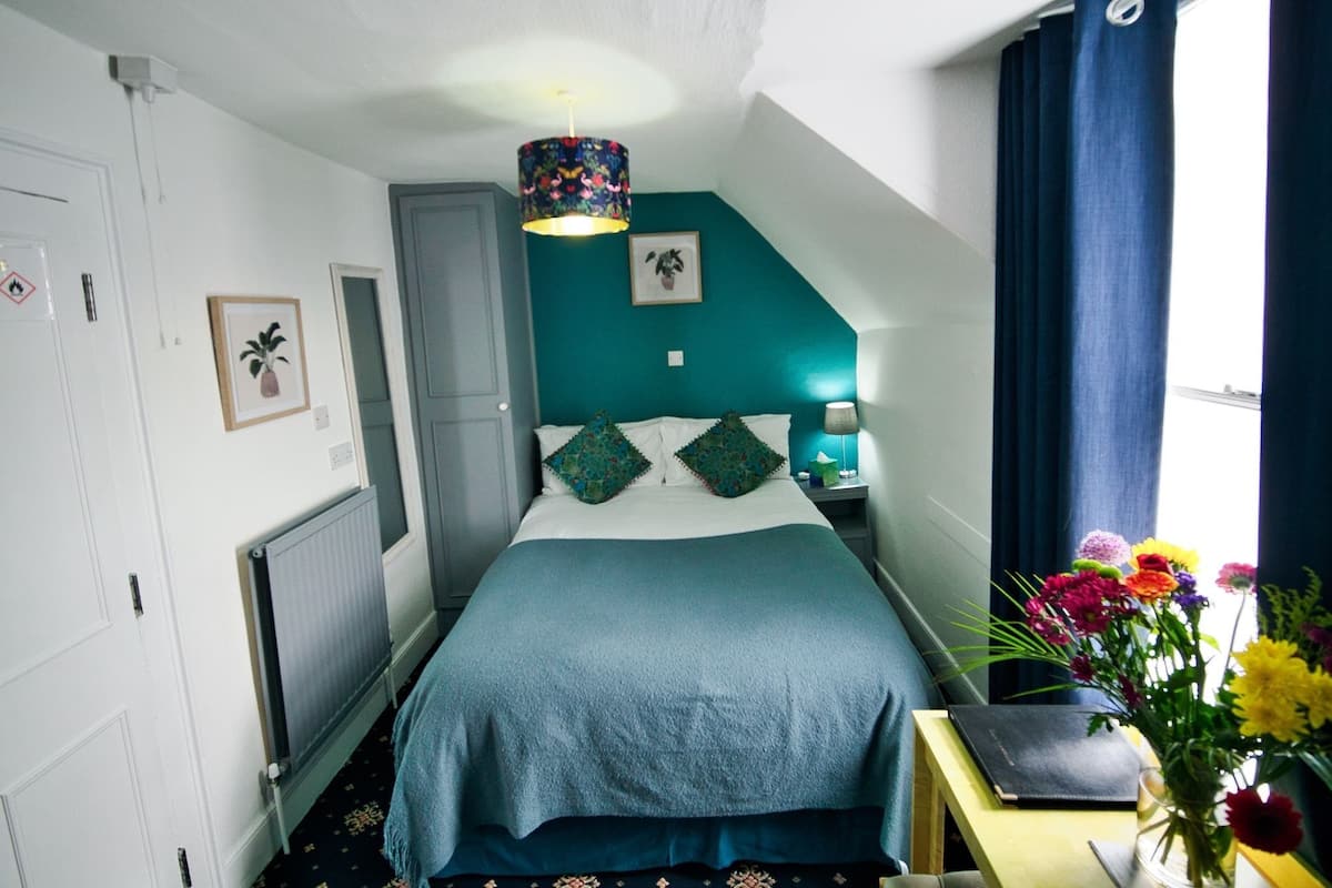 Um quarto com cama, mesa de cabeceira, escrivaninha, cadeira, janela com cortinas, radiador e um armário com porta e um quadro.