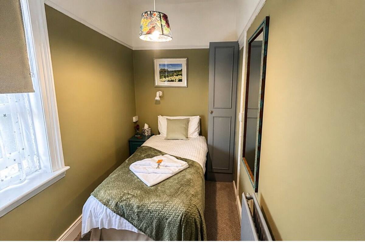 Um quarto pequeno e acolhedor com uma cama, uma mesinha de cabeceira, uma luminária e uma janela com cortinas.