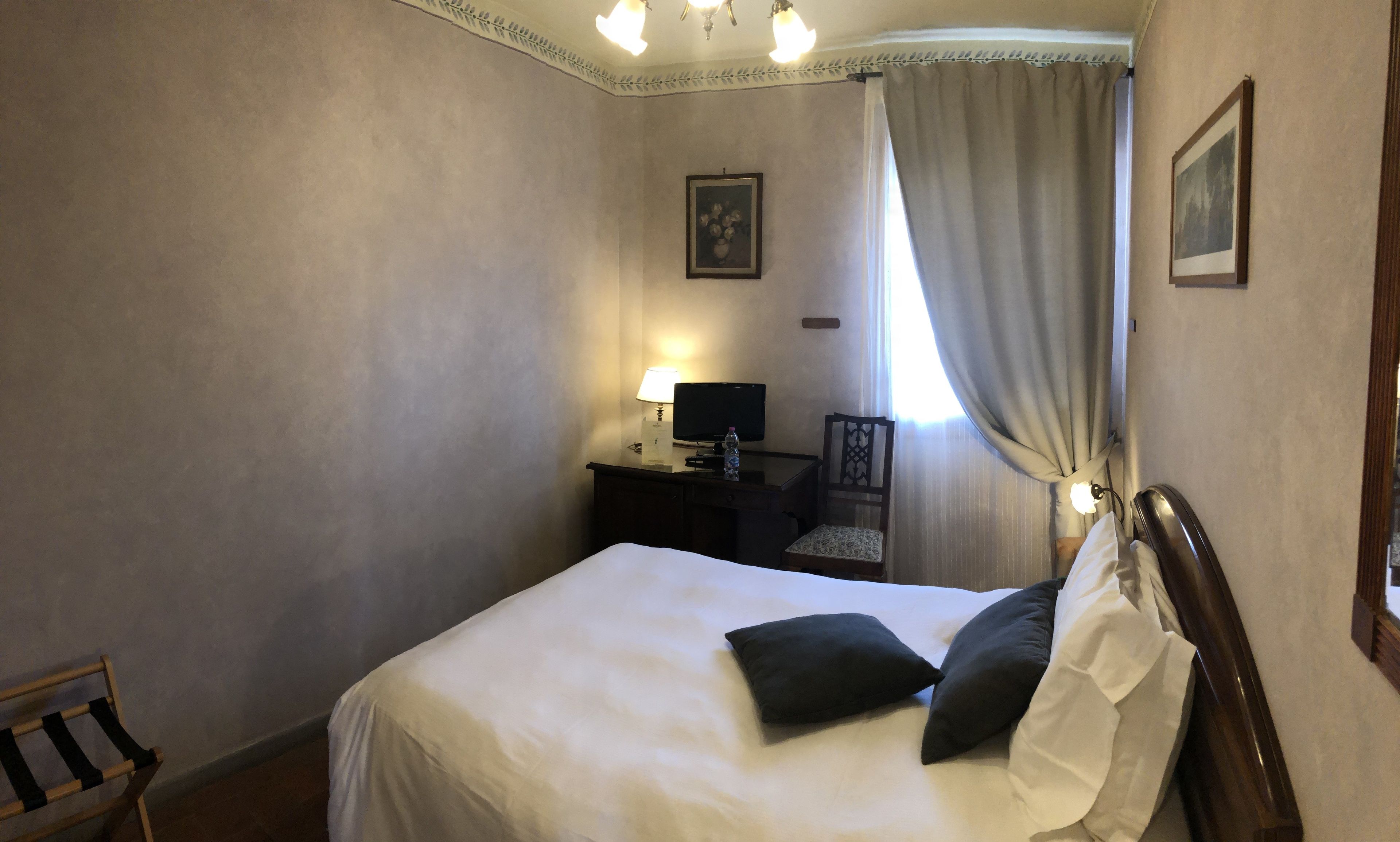 Quarto de hotel com cama, escrivaninha com luminária, cadeira e janela com cortinas.