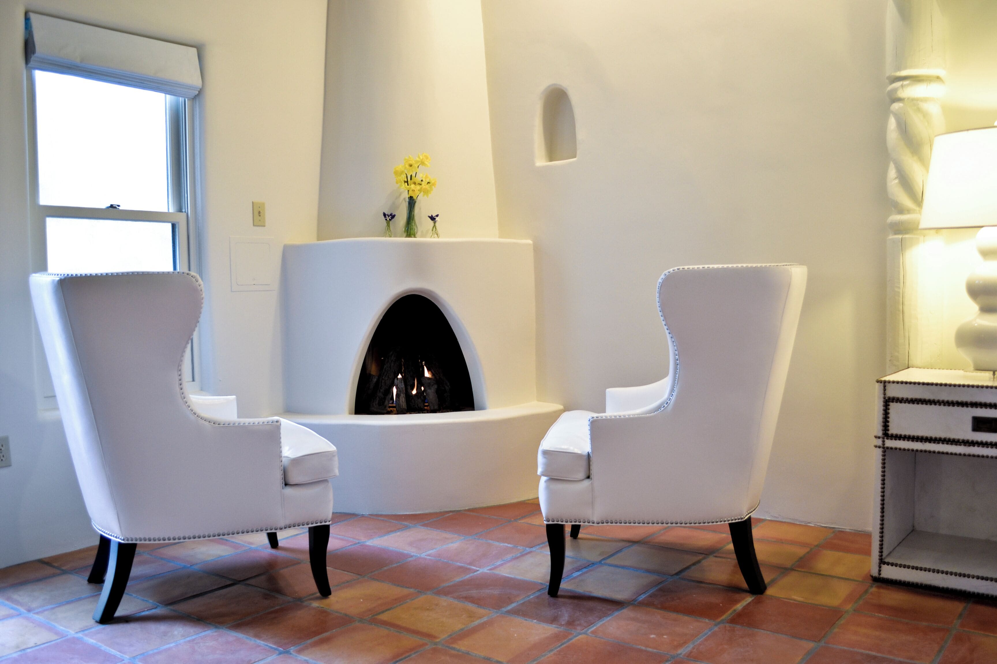 Quarto com duas poltronas brancas, lareira, janela e abajur.