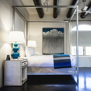 Quarto moderno com cama de dossel, duas luminárias de cabeceira, um quadro de nuvens e vista para o exterior através de uma grande janela.
