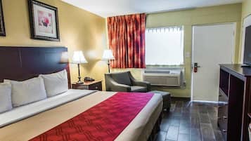 Quarto de hotel com uma cama grande, duas luminárias de cabeceira, uma cadeira, uma escrivaninha, uma televisão e uma porta.