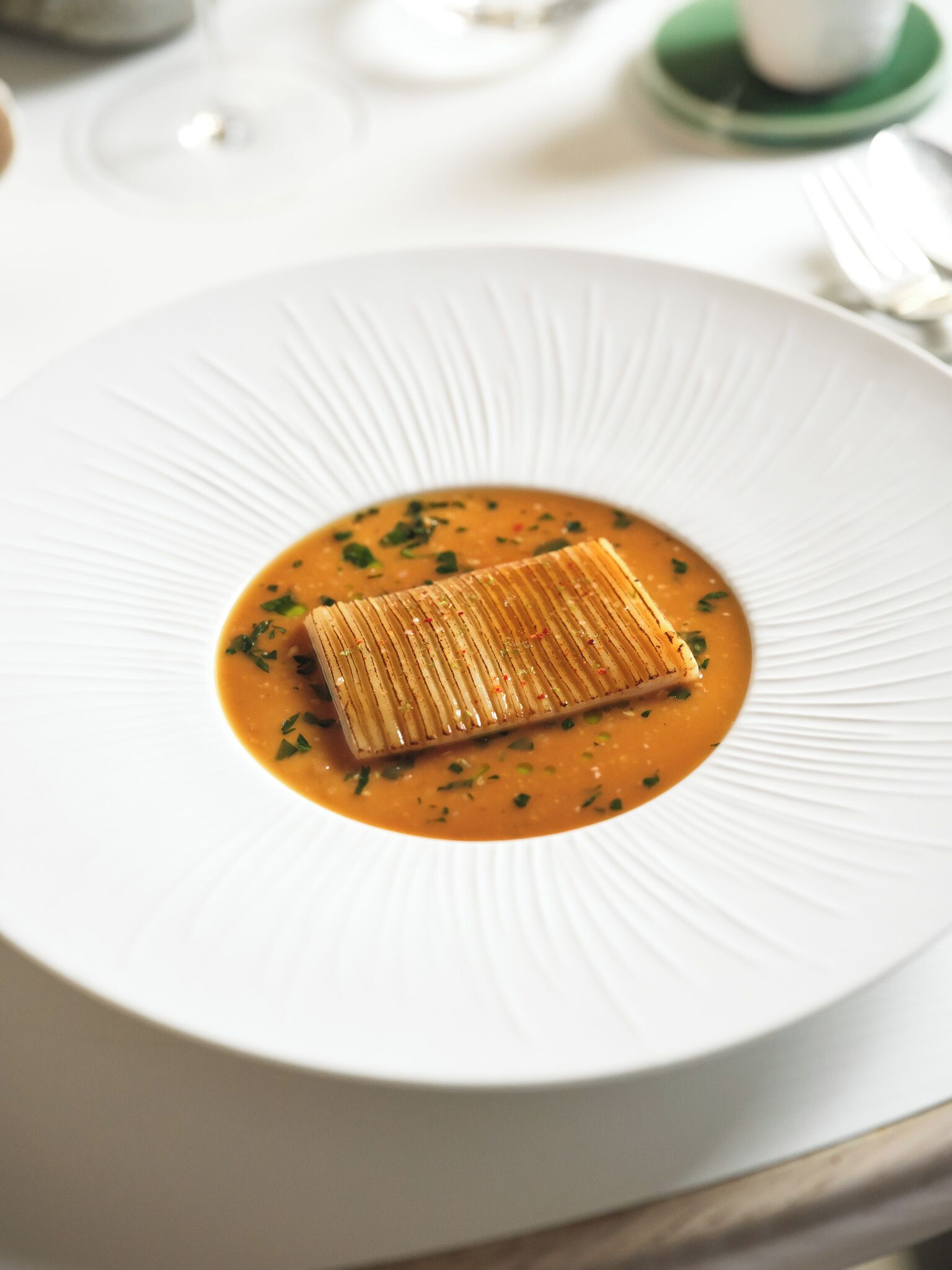 Um prato branco com um pedaço retangular de comida, possivelmente um filé de peixe, sobre um leito de molho laranja, guarnecido com ervas verdes.