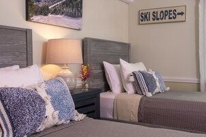 Quarto de hotel com duas camas, mesa de cabeceira com luminária, um vaso com flores e um aviso na parede que diz 'SKI SLOPES'.